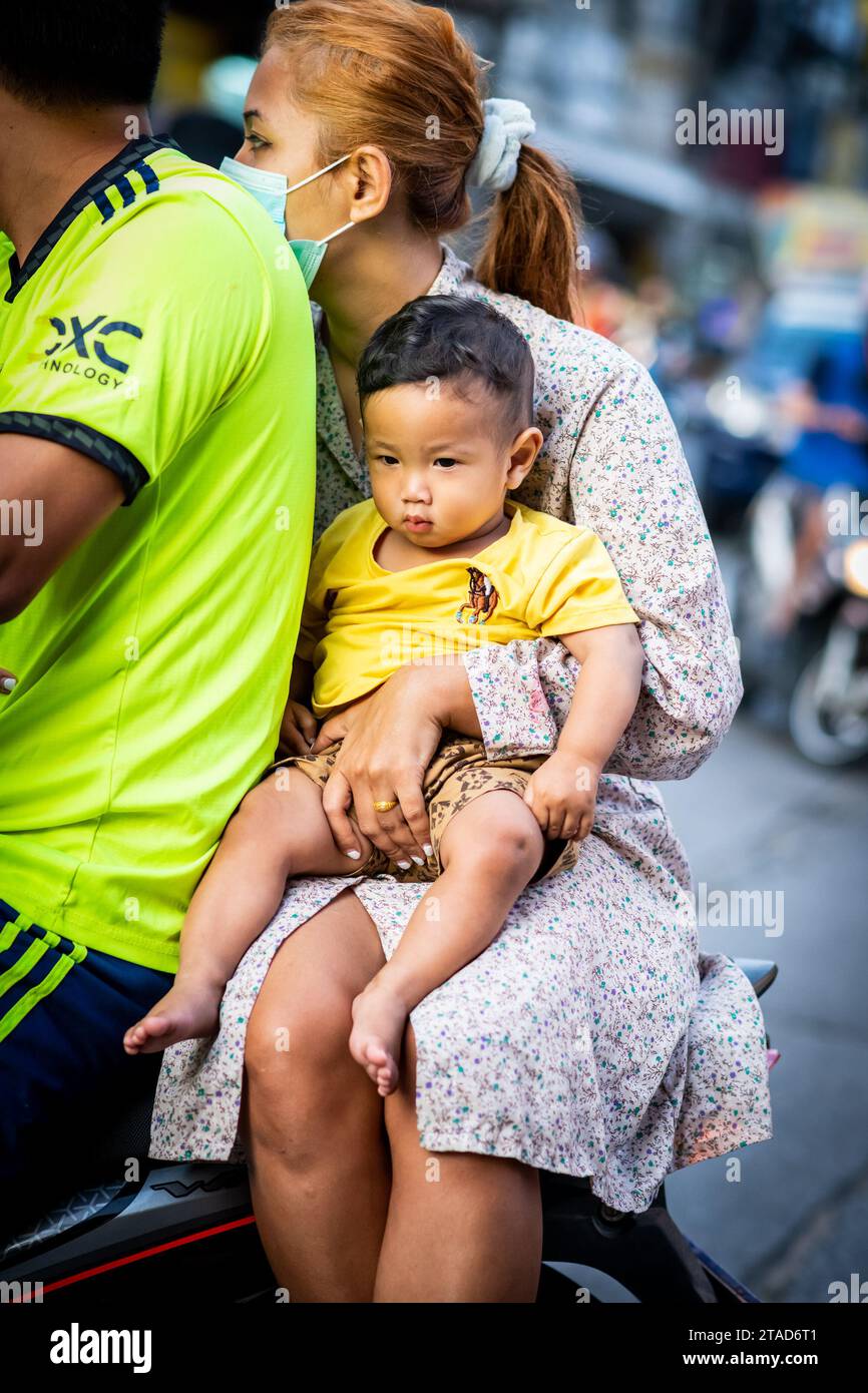 A little Thai boy is sat on his mums lap at the back of a motorbike taxi travelling through the ...
