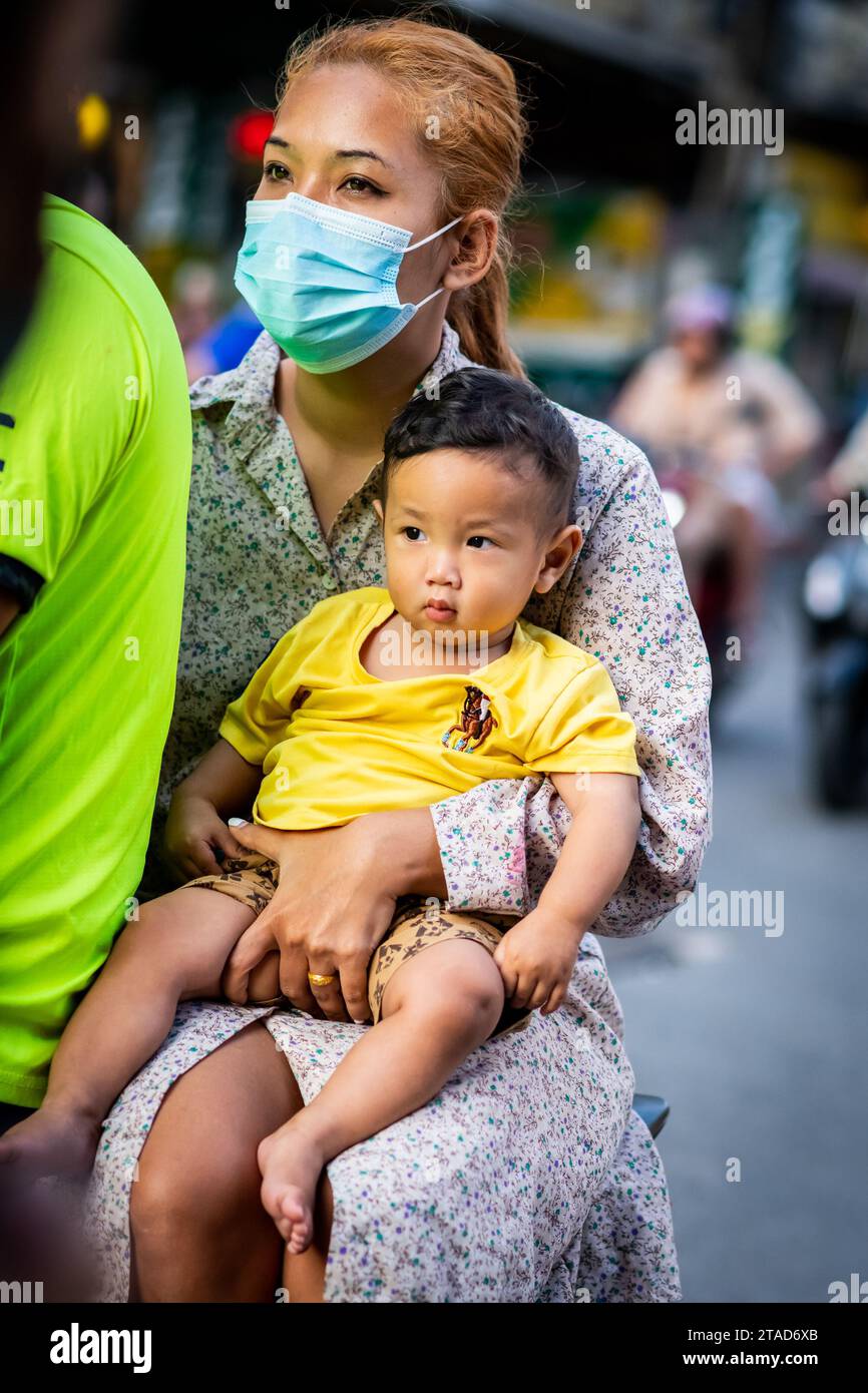 A little Thai boy is sat on his mums lap at the back of a motorbike ...