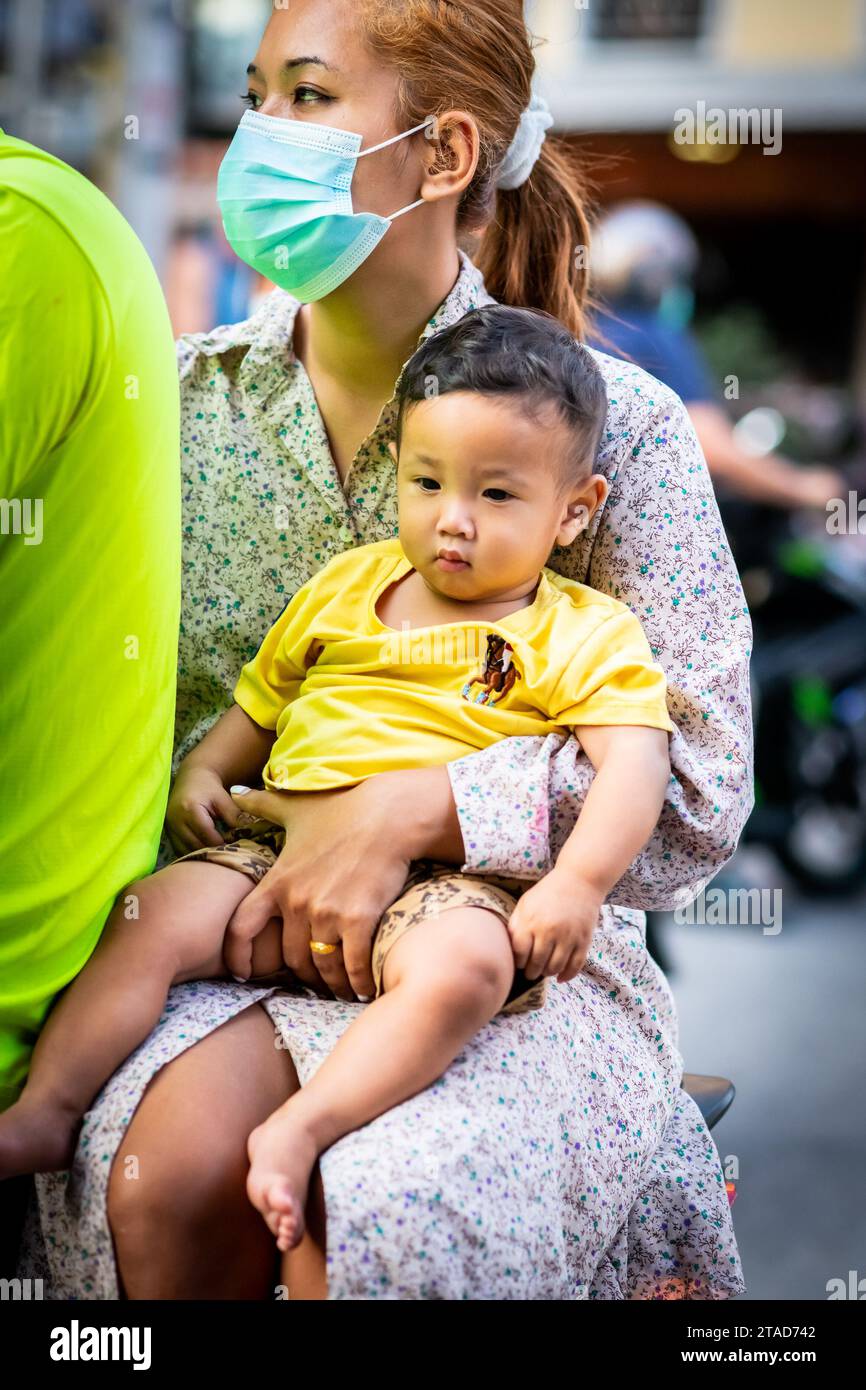 A little Thai boy is sat on his mums lap at the back of a motorbike ...