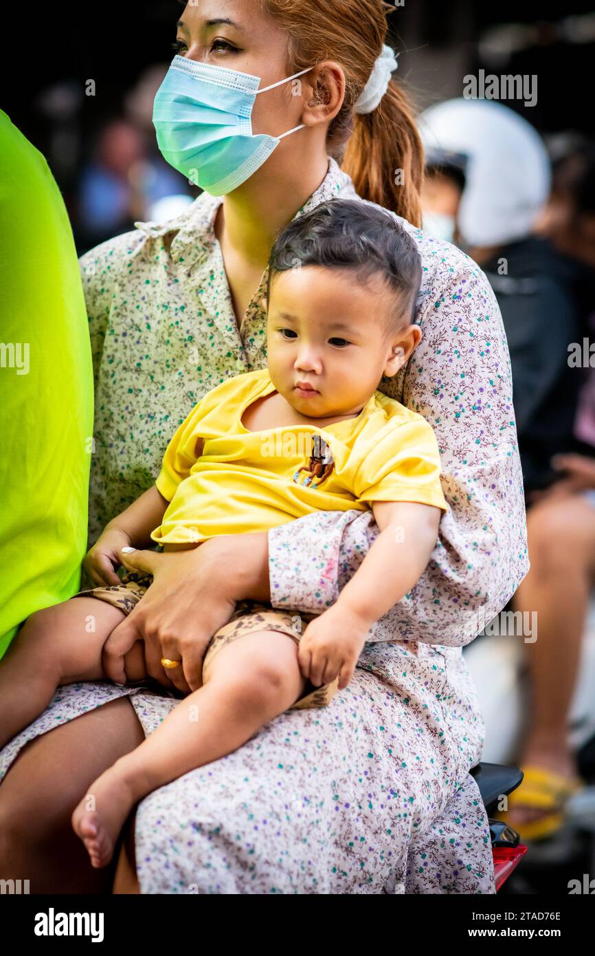 A little Thai boy is sat on his mums lap at the back of a motorbike ...