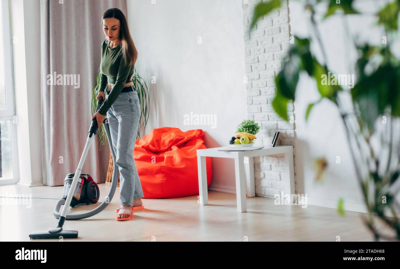 Full-length photo of beautiful young woman is using a vacuum cleaner ...