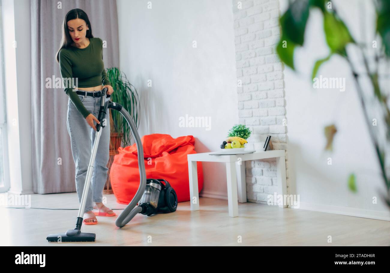 Full-length photo of beautiful young woman is using a vacuum cleaner ...