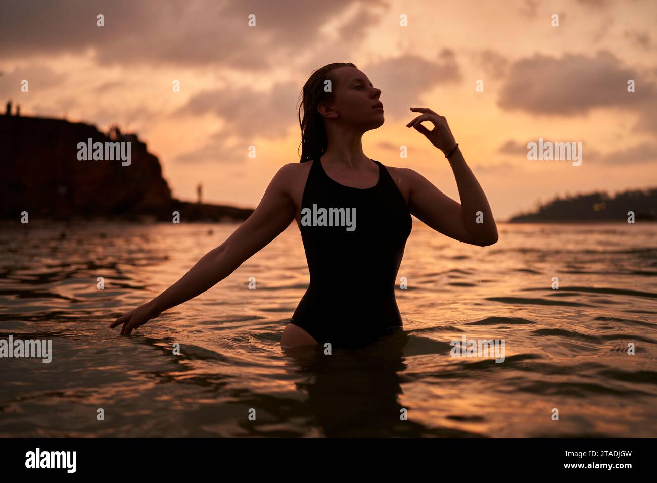 Silhouetted woman stands in sea at dusk, touching chin thoughtfully ...