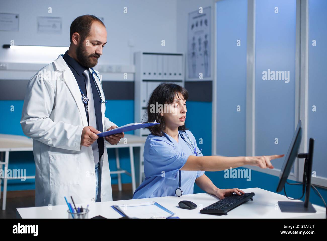 Caucasian doctor and female nurse in blue scrubs and lab coat collaborate in clinic office ...