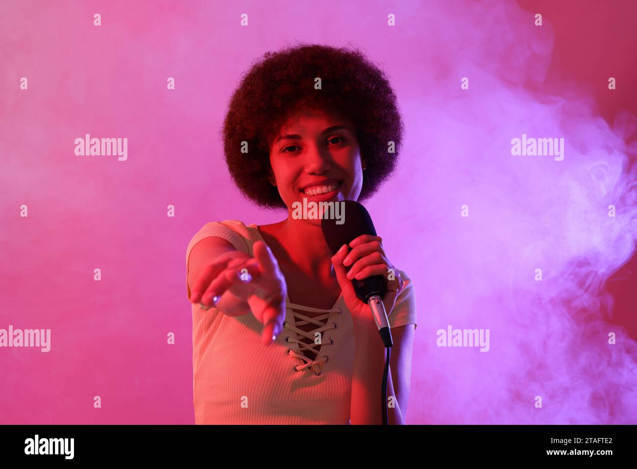 Curly young woman with microphone singing on pink background. Color ...