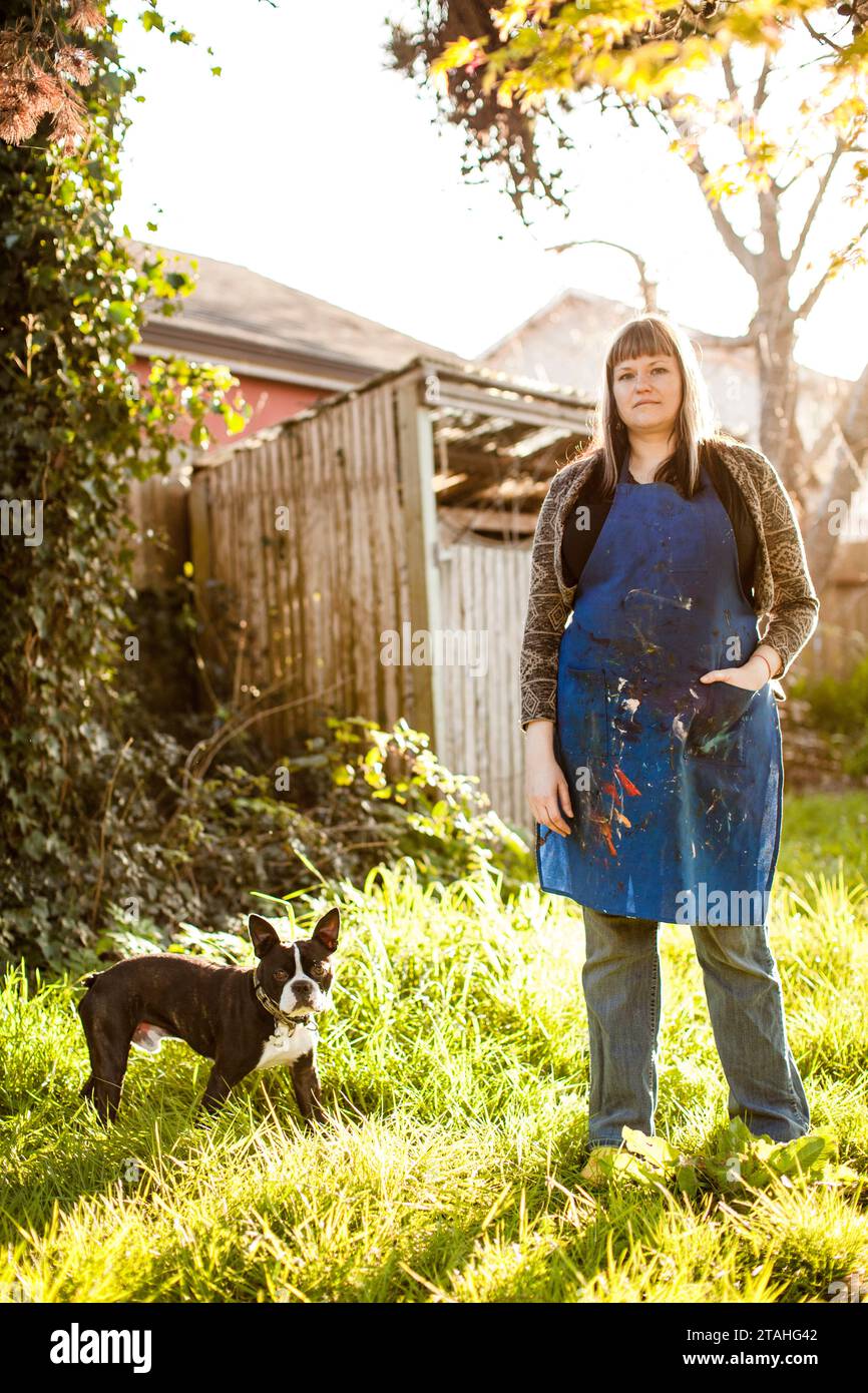 Female Artist in messy apron with small dog backlit in grassy yard ...