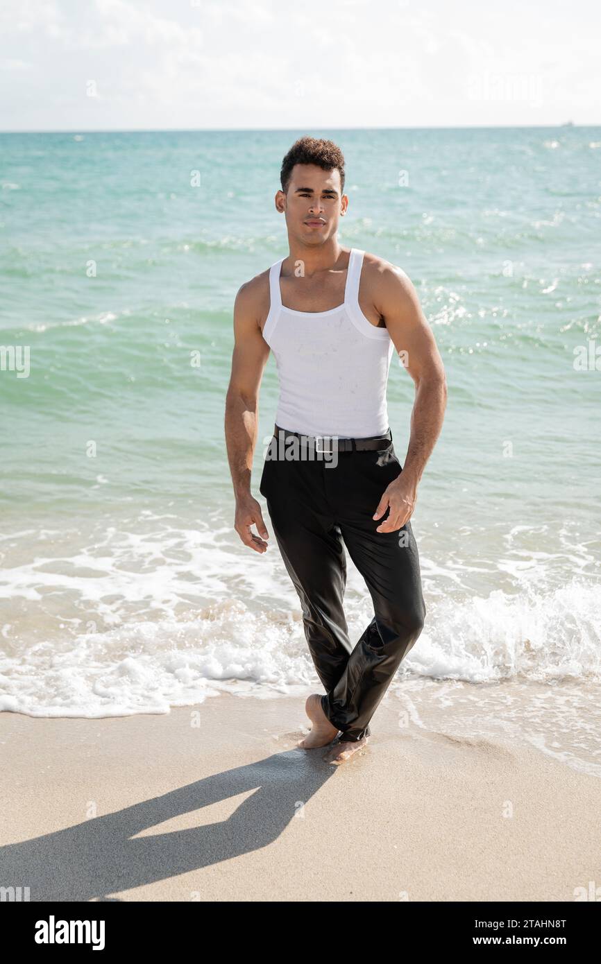 cuban man looking at camera while standing on sand near ocean water in ...