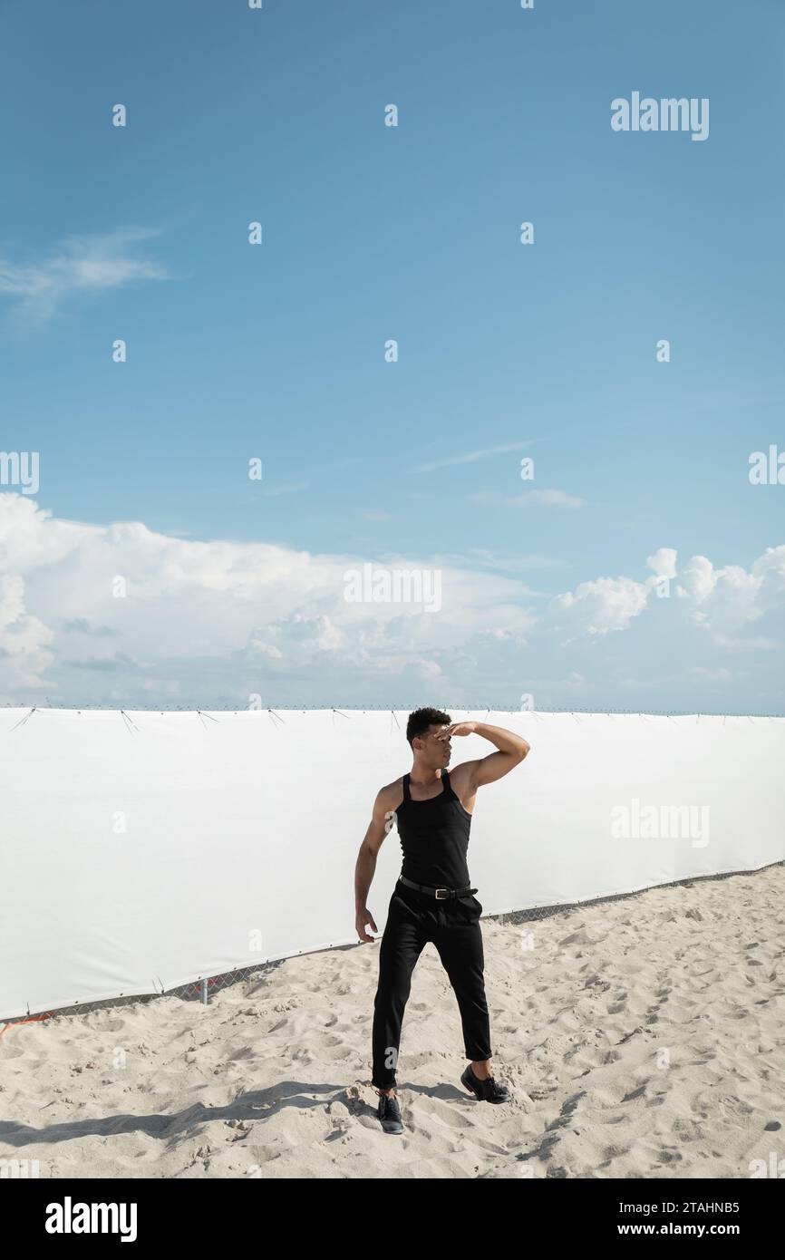 Muscular and young cuban man in stylish outfit covering face from ...