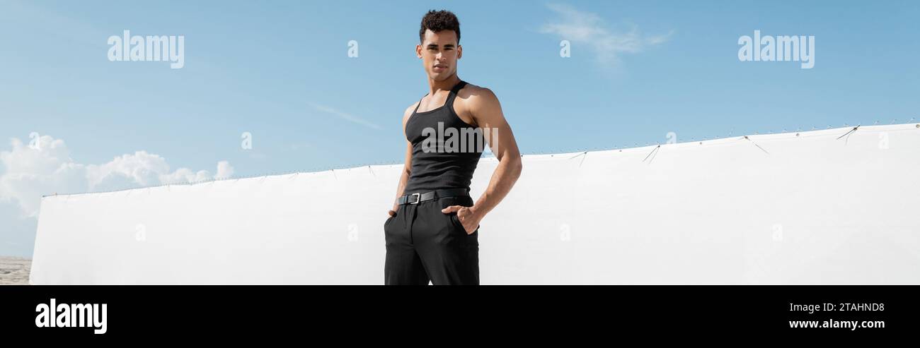 muscular young cuban man in black attire posing with hands in pockets ...