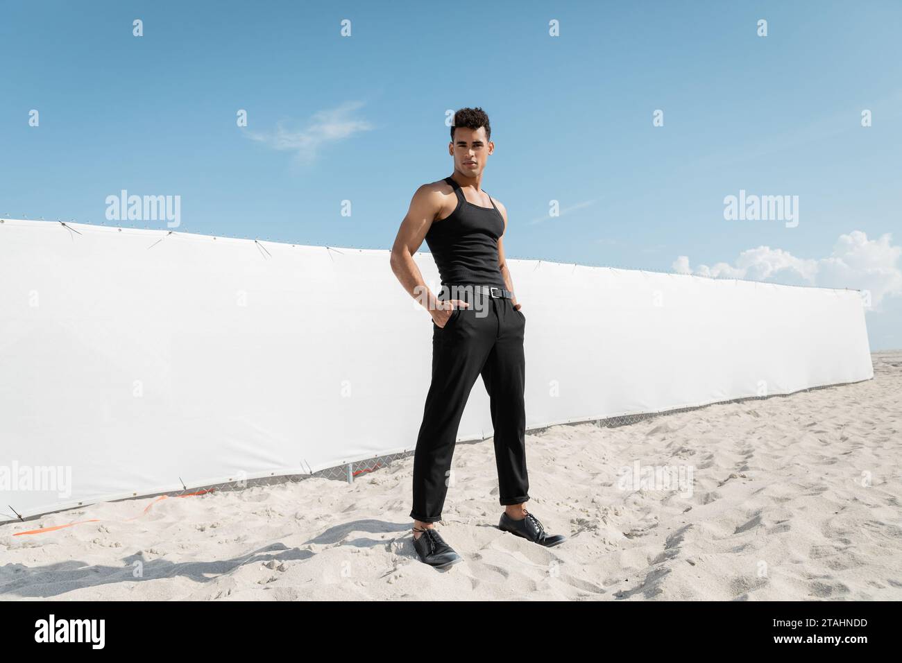 muscular young cuban man in black attire posing with hands in pockets ...