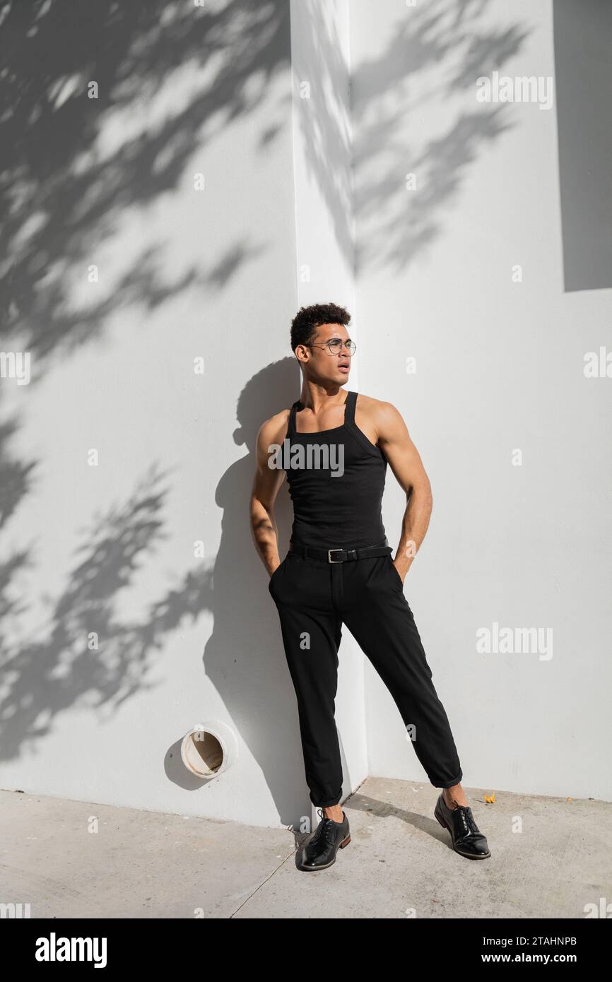 stylish young cuban man posing near white wall with shadow on urban ...