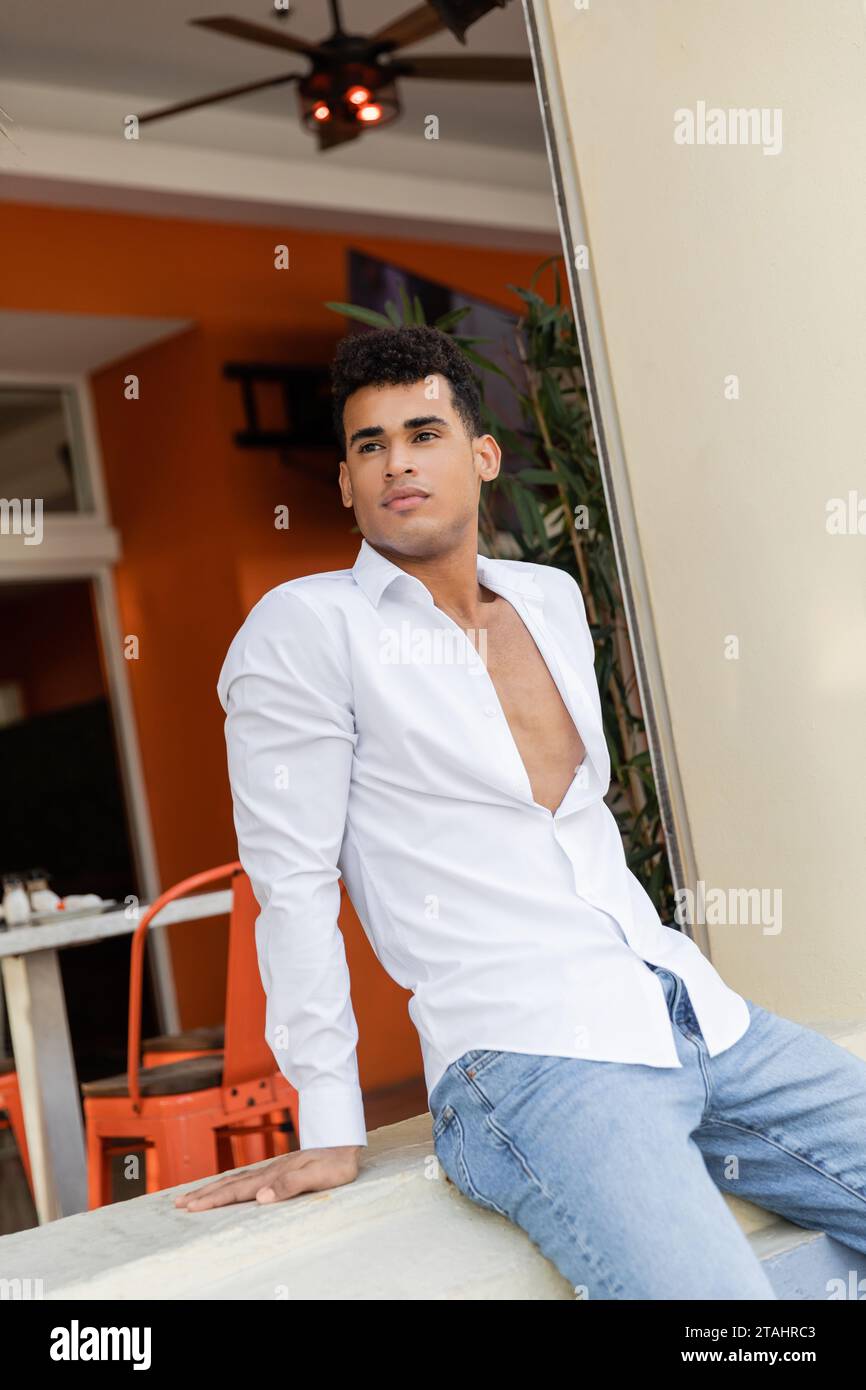 curly cuban man in white shirt and jeans sitting on parapet of outdoor ...