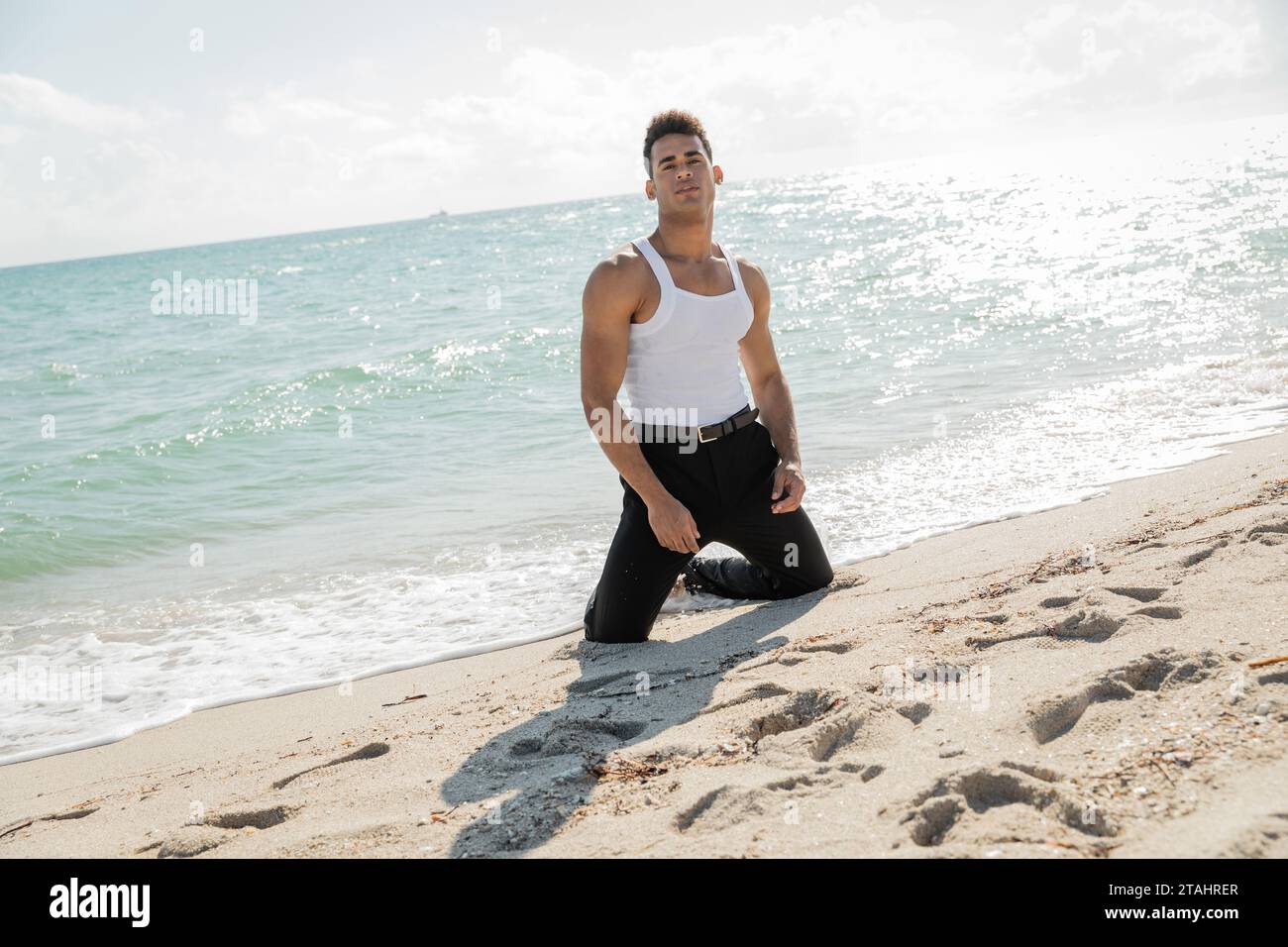 young cuban man in stylish clothes posing on coast sand near ocean and ...