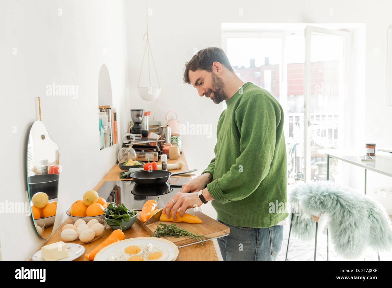 smiling man in green jumper cutting fresh pepper while cooking ...