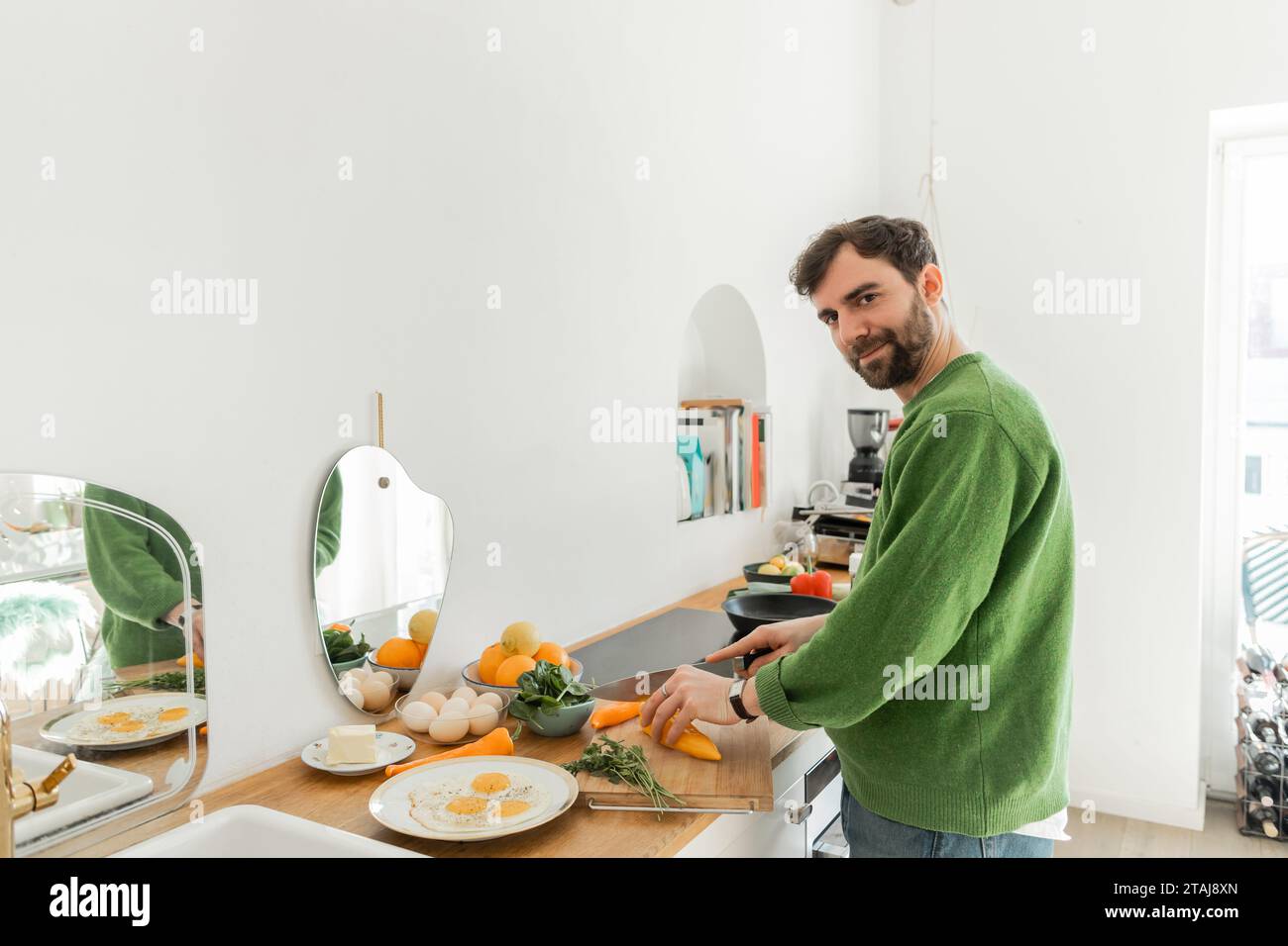 happy man in green jumper cutting fresh pepper while cooking breakfast ...