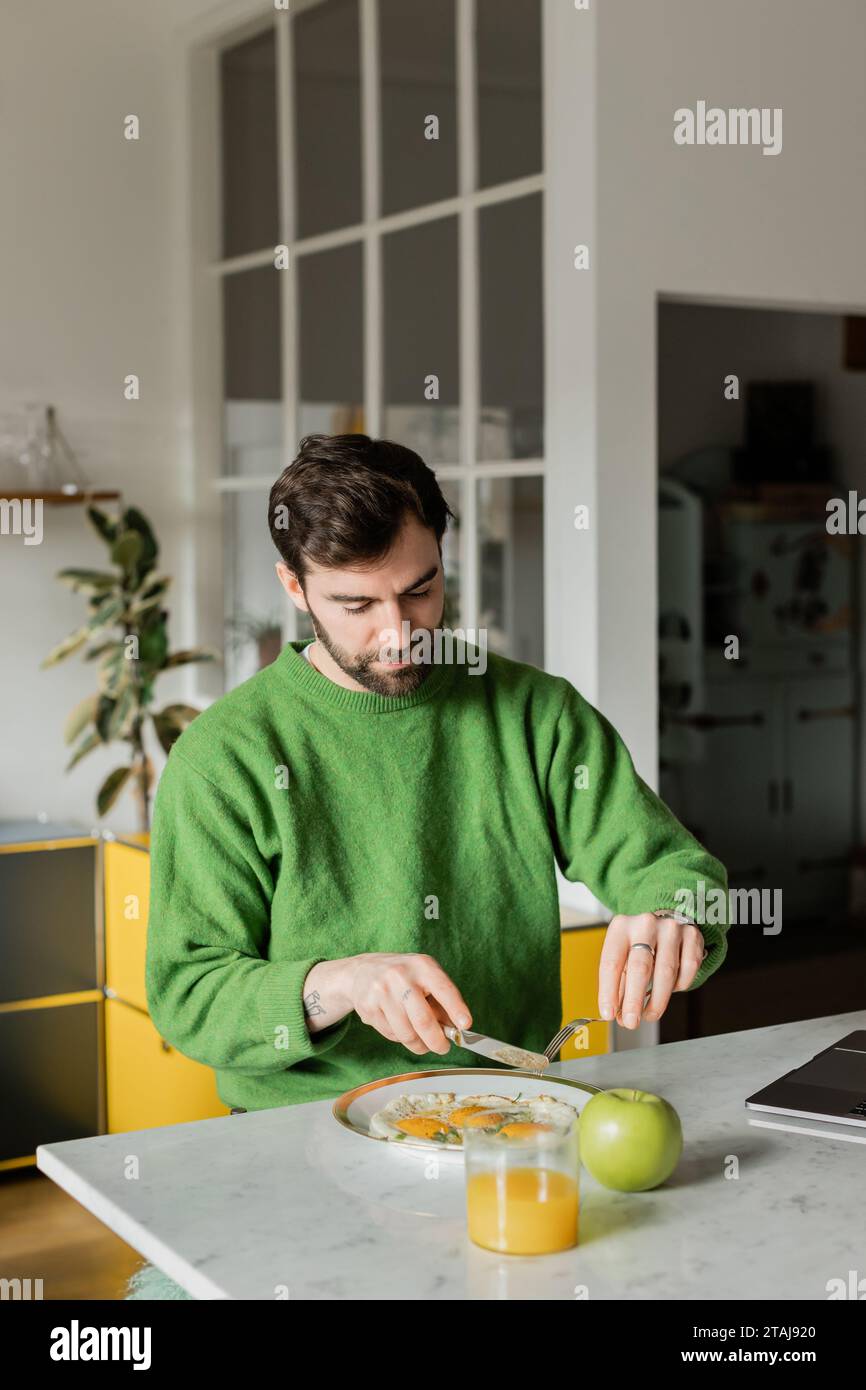 bearded man in green jumper holding cutlery near breakfast in modern ...