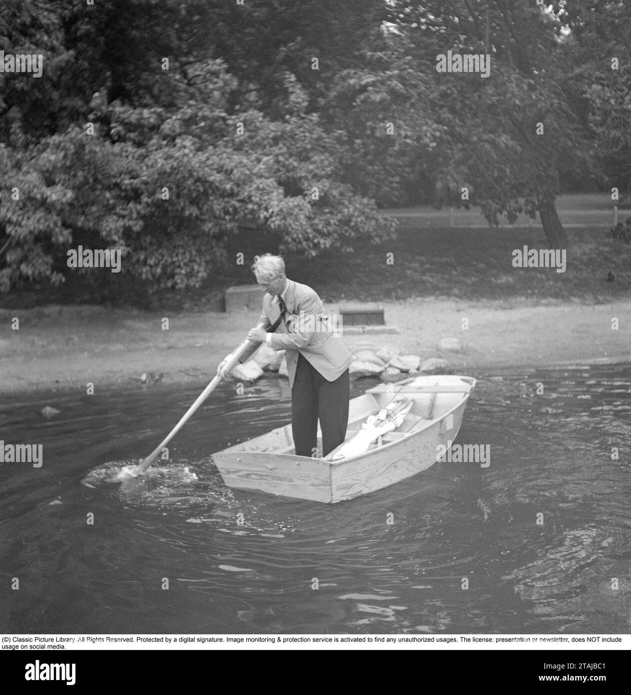 In the 1940s. A man standing up in a small sailing boat, using an oar ...