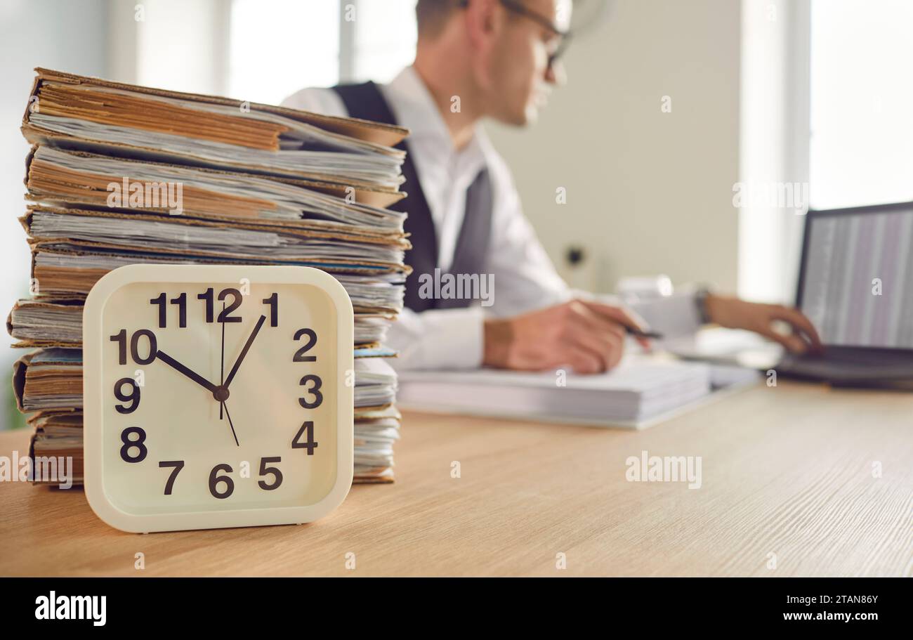 Clock and stack of paperwork on office table, with man working on ...