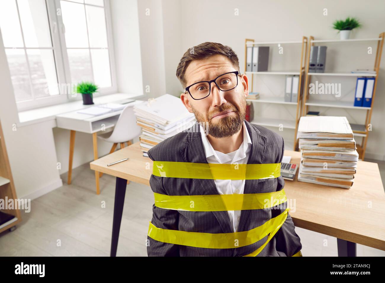 Funny office slave, wrapped with adhesive tape, sitting by desk with ...
