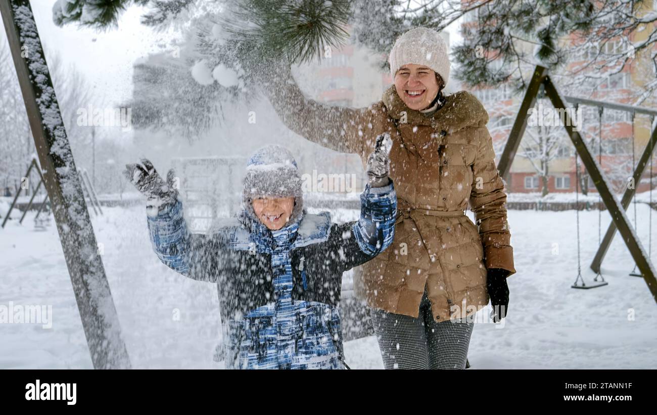 Laughing mother with son shaking tree branch and enjoying falling snow ...