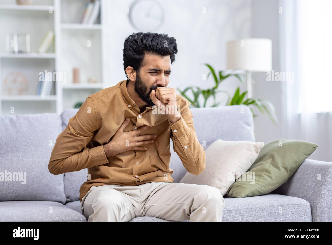 Indian young man sitting at home on the couch and coughing, covering ...