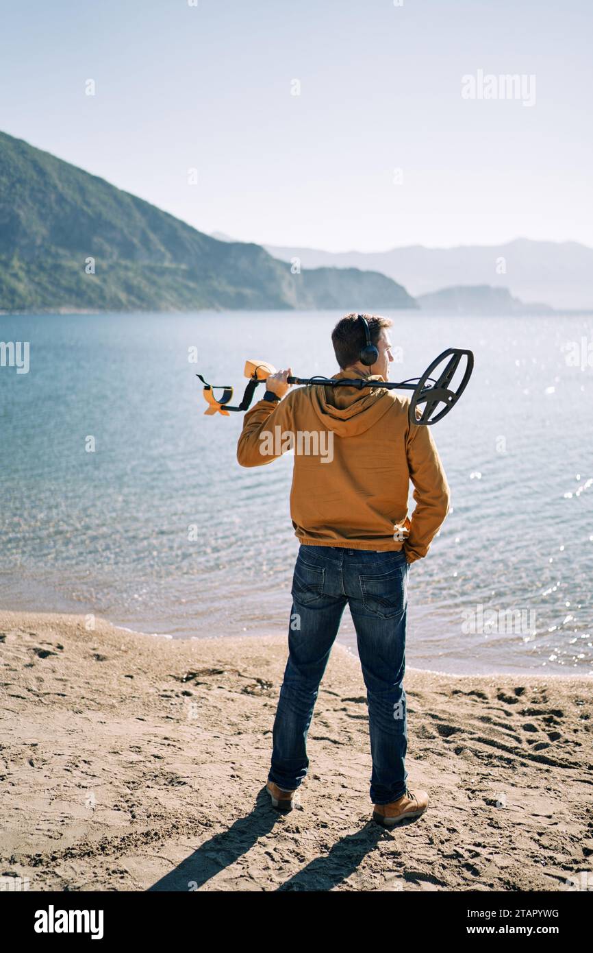 Beach cop stands with a metal detector on his shoulder and looks into ...