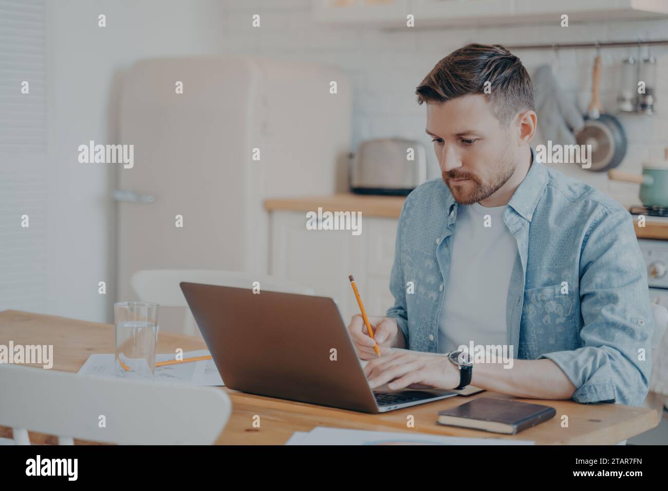 Concentrated man working on laptop with notes, in a well-lit kitchen ...