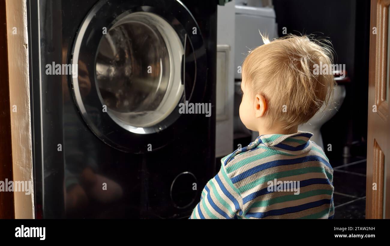 Cute baby boy sitting in bathroom and looking at working washing ...