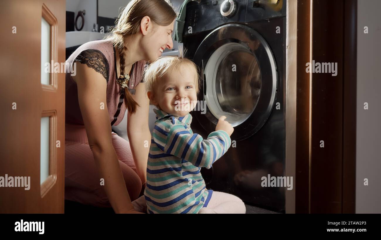 Happy mother with baby boy doing laundry and looking at washing machine ...