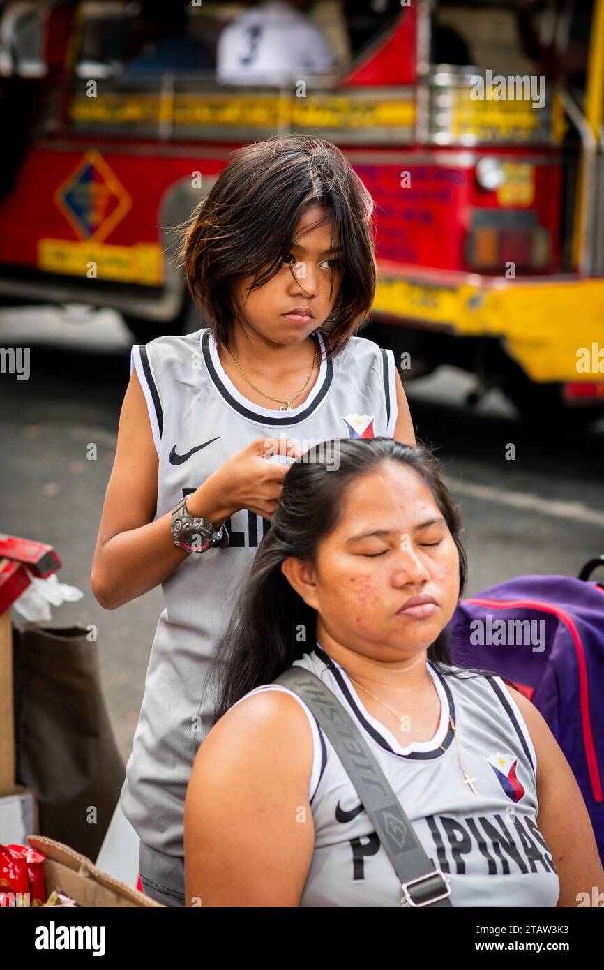 A young filipino girl cleans her mothers hair sat at their street stall ...