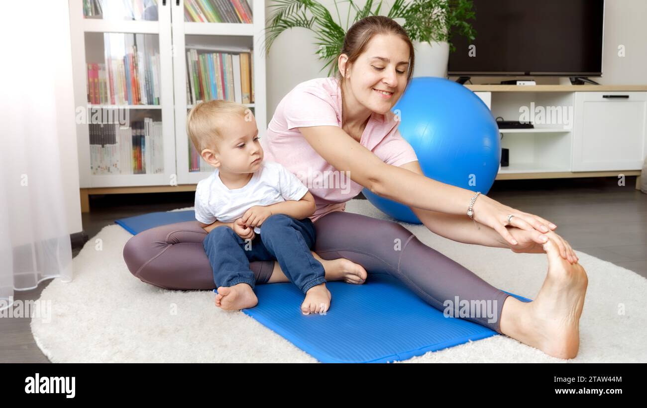 Cute baby boy looking at mother stretching leg muscles on yoga mat in ...