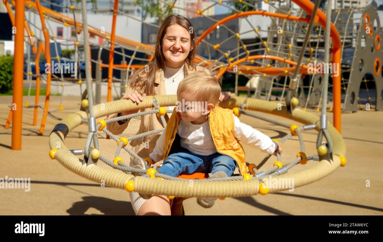 Happy mother pushing swing with her baby son on playground. Children ...