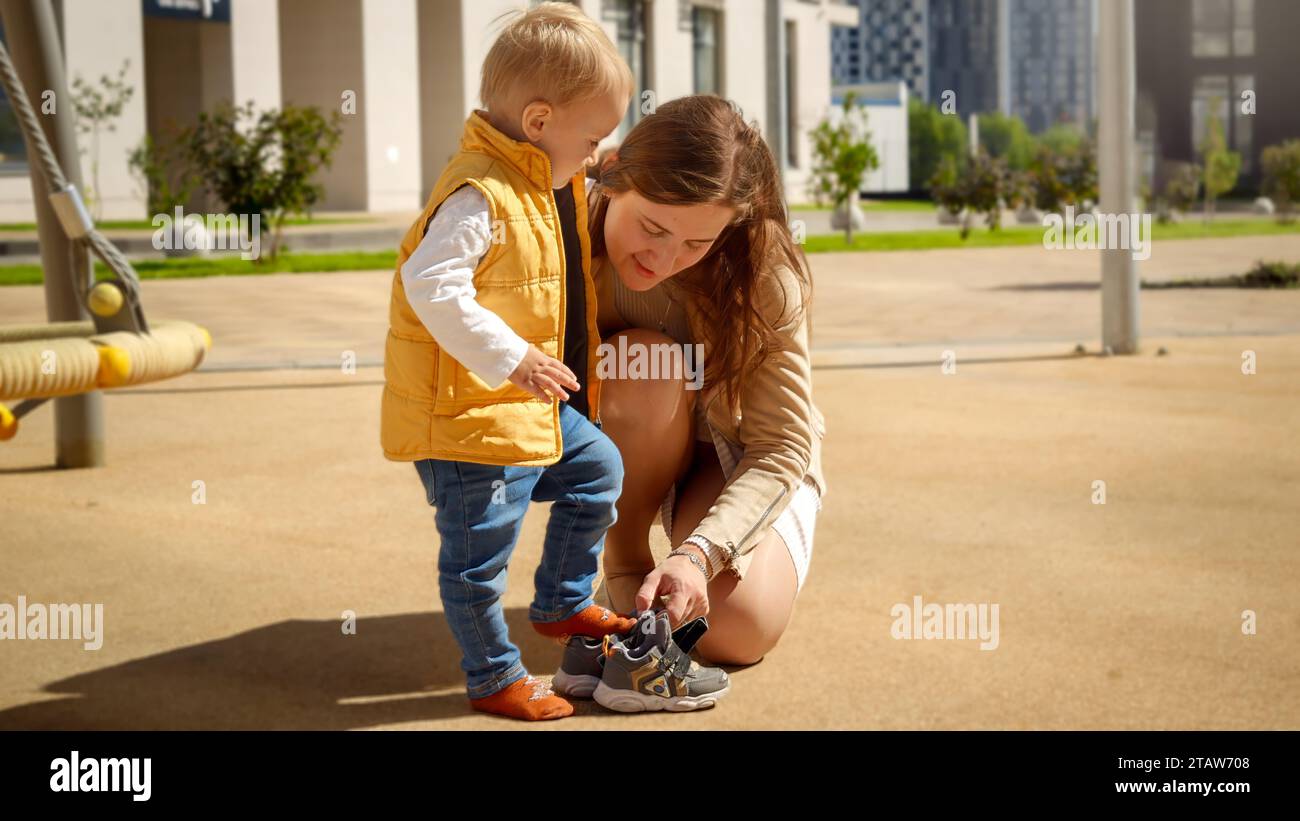 Young caring mother helping her toddler son to put on shoes on ...