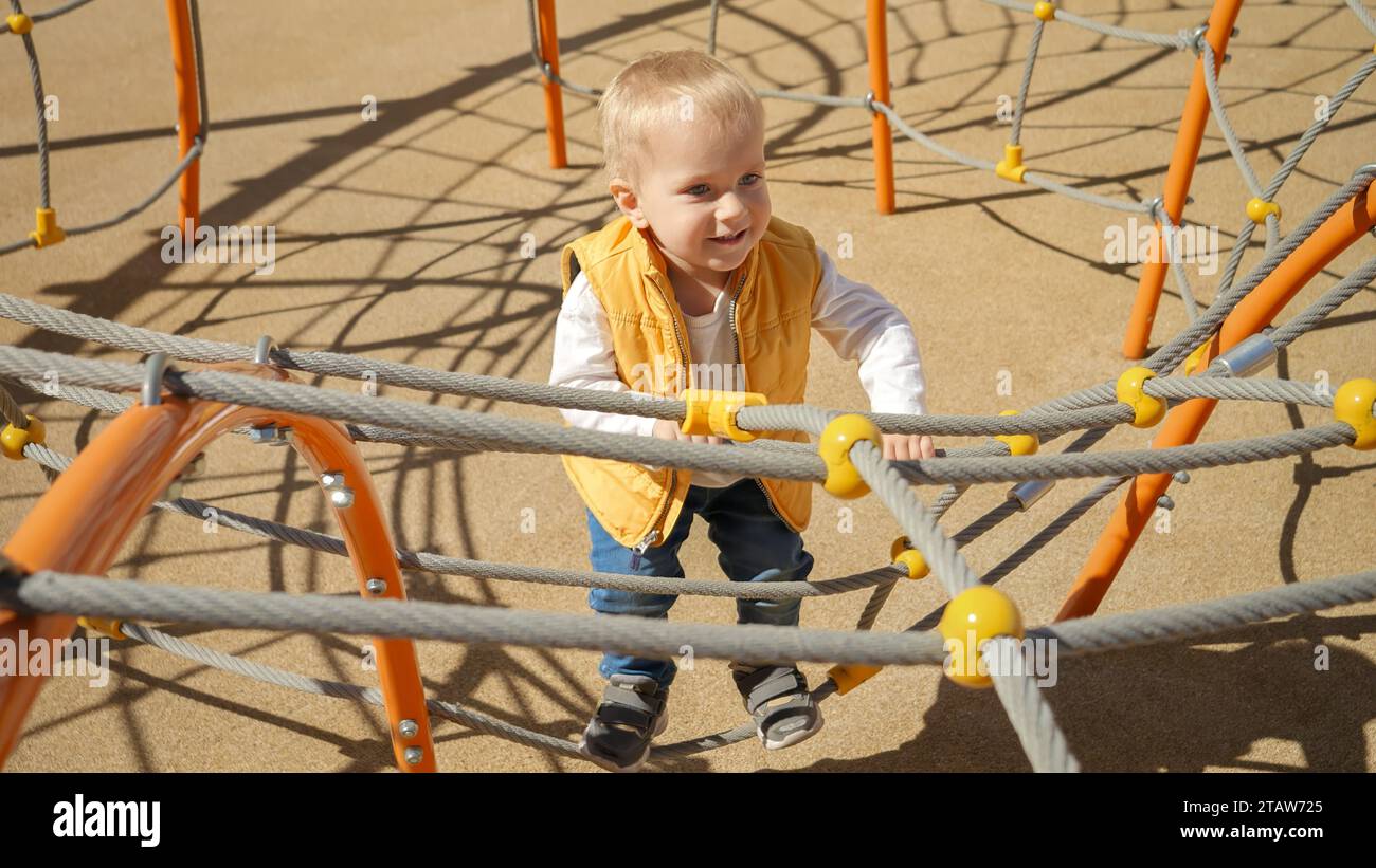 Cute baby boy playing on the playground and climbing up the rope spider ...