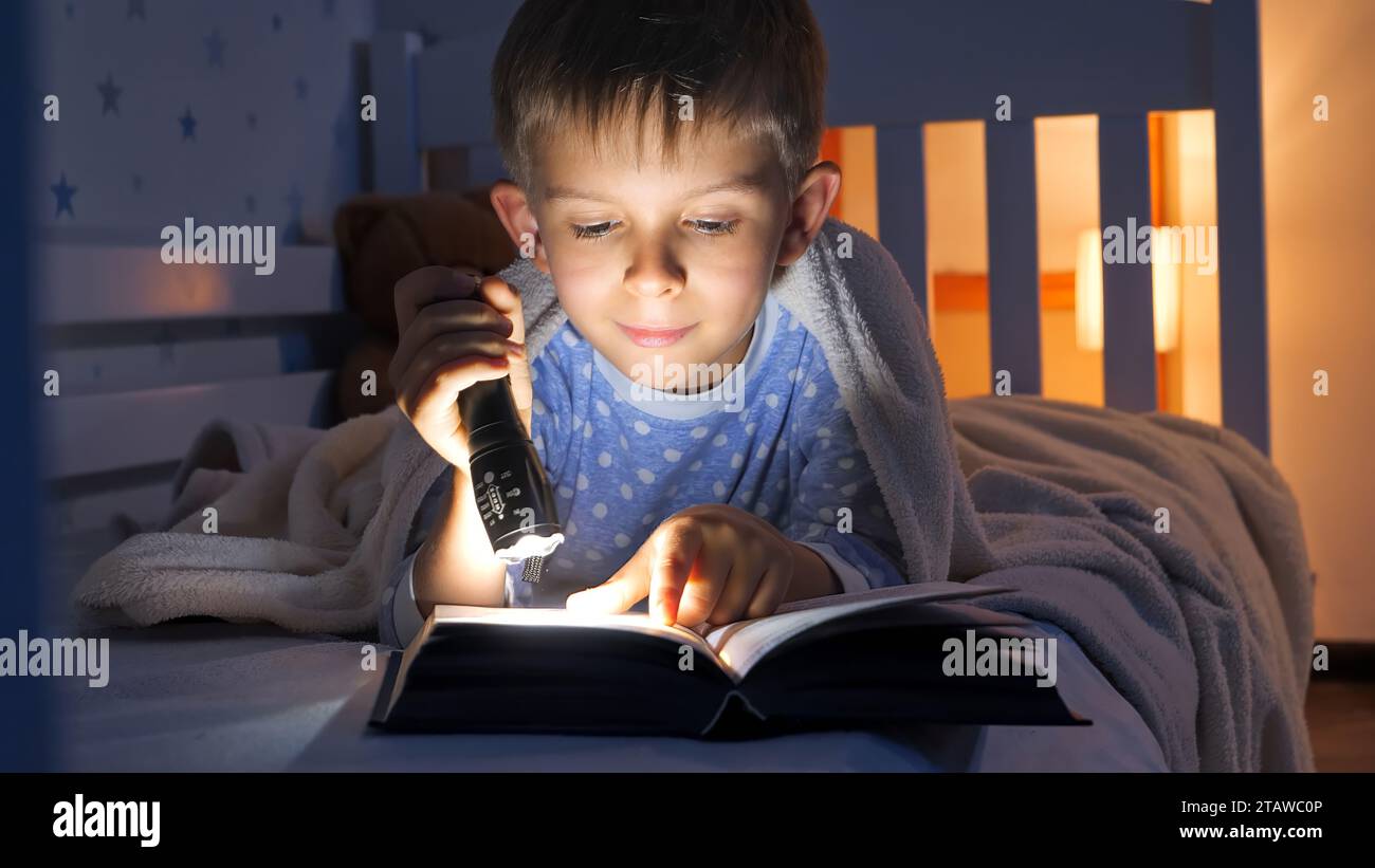 Little boy in pajamas lying in bed and reading bedtime story book with ...