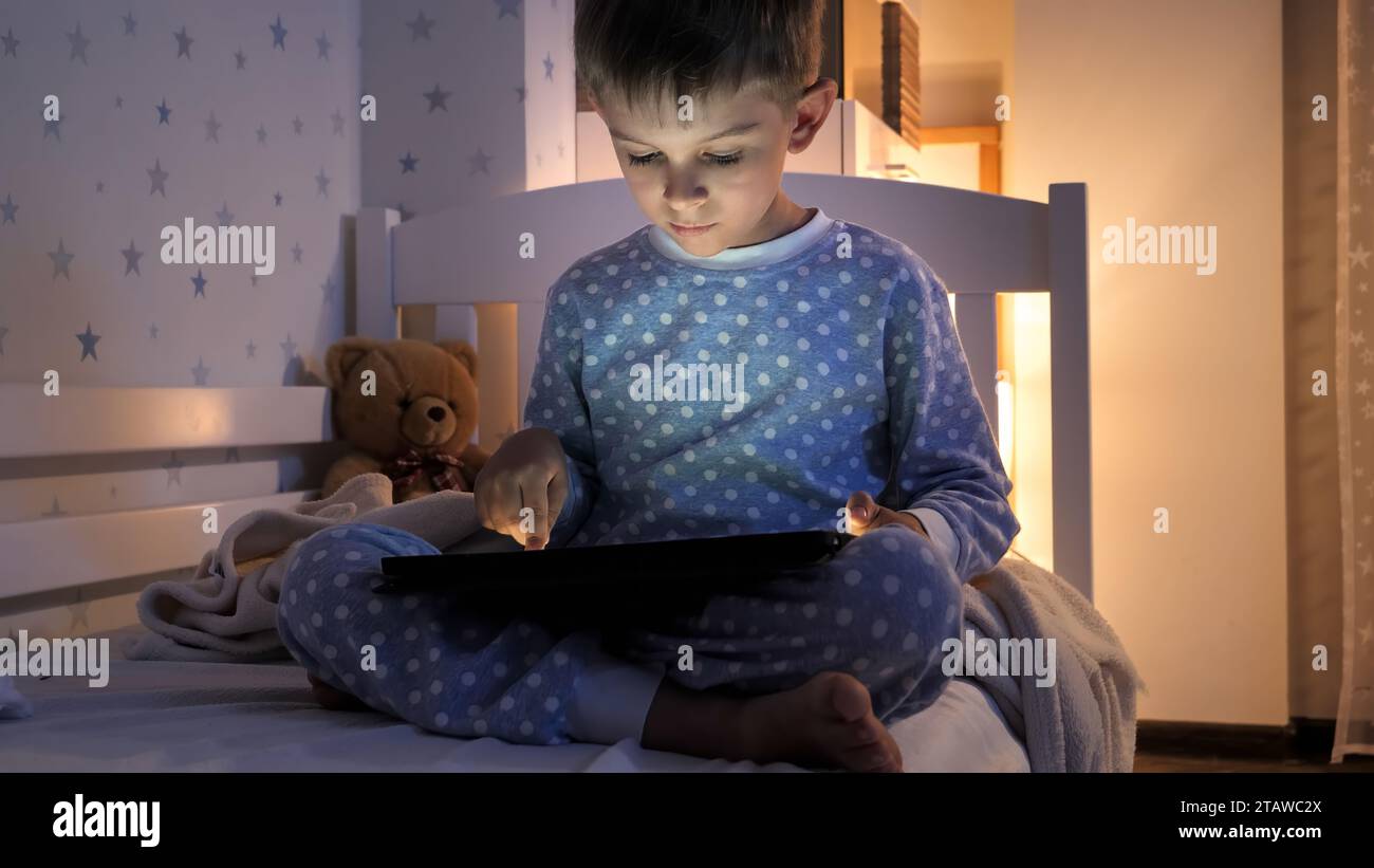 Little boy siting on bed at night and watching cartoons on tablet ...