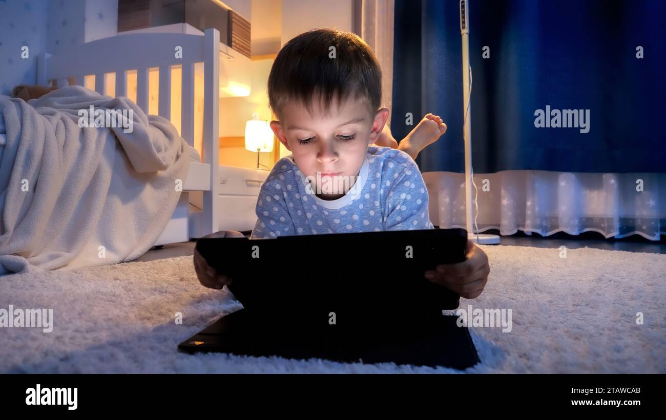 Cute boy in pajamas lying on floor in his room and using tablet ...