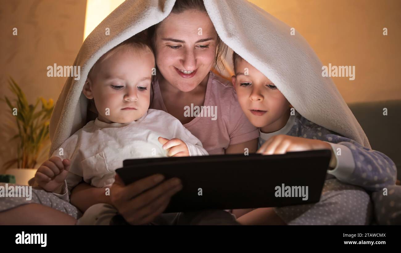 Young mother with two sons covering under blanket and watching video on ...
