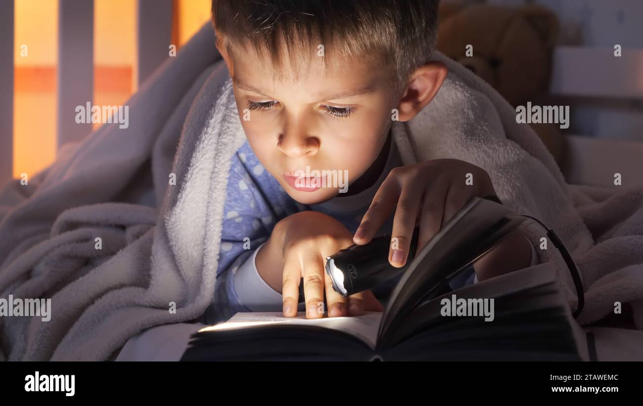 Closeup of boy holding flashlight reading book in bed. Children ...