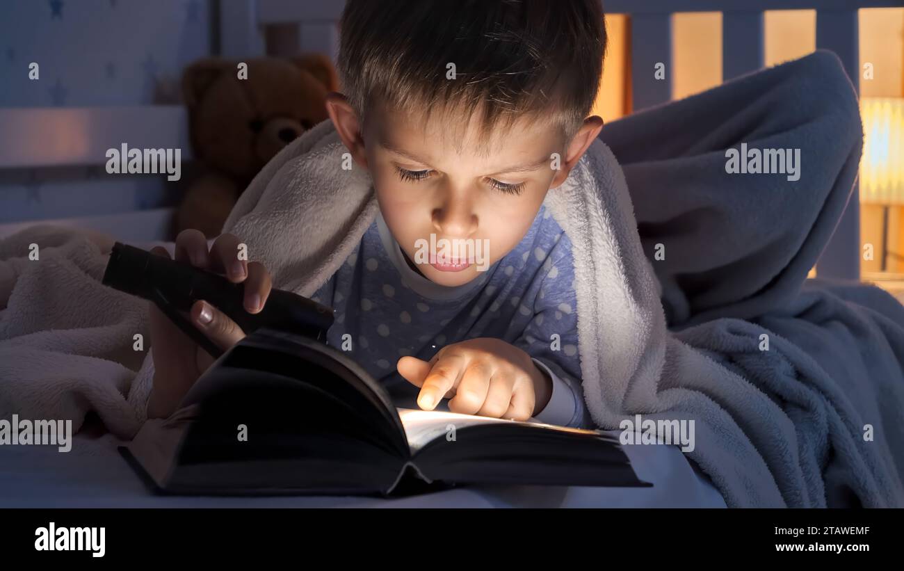 Portrait of boy with torch lying in bed and reading story book ...