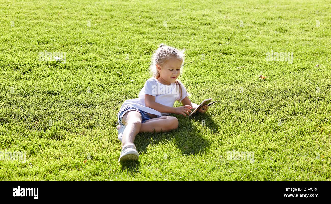 Little girl sitting on the lawn in the summer city park and holding smartphone. Childhood ...
