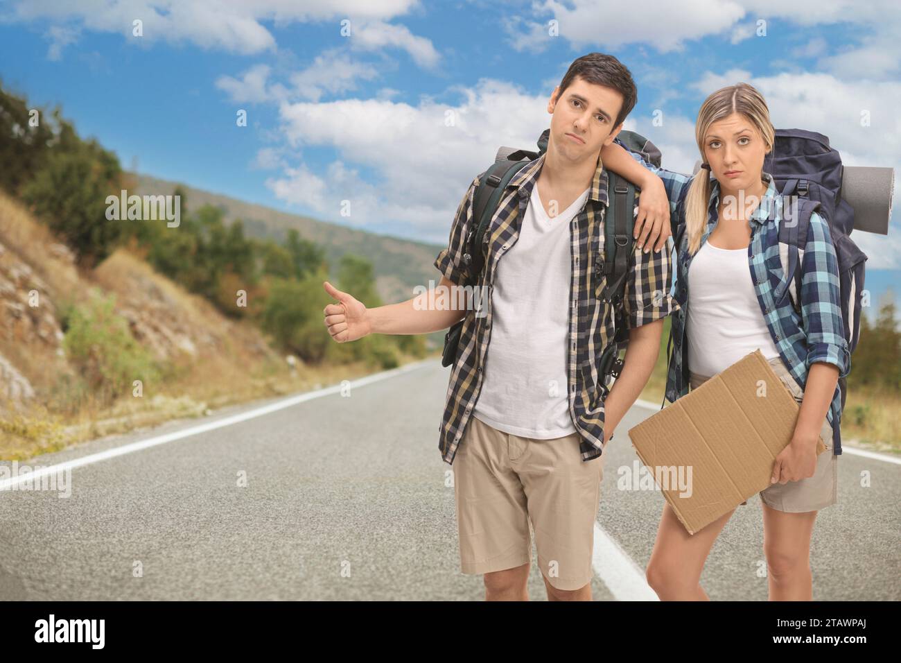 Sad hikers holding a blank cardboard sign and hitchhiking on a road ...