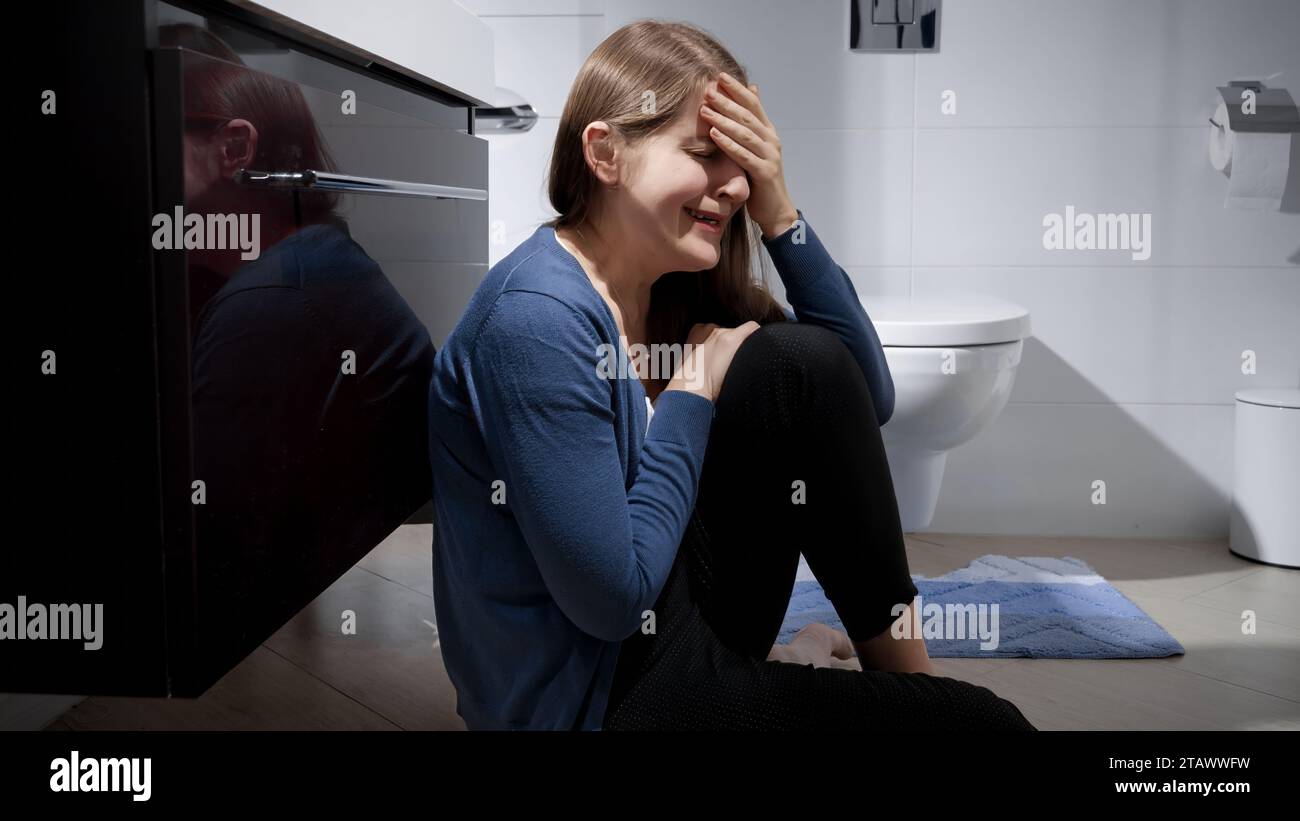 Crying woman sitting on floor in bathroom and leaning on sink. Concept ...
