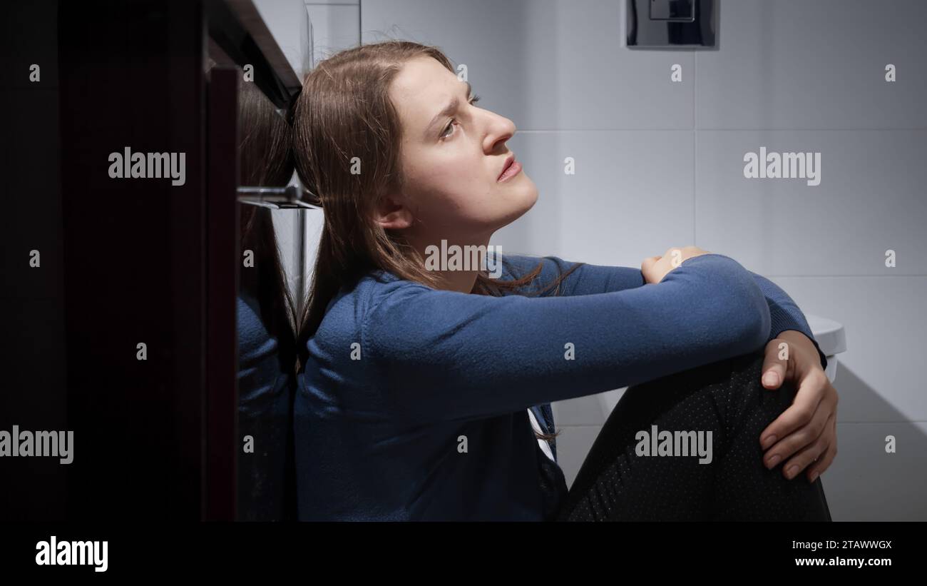 Portrait of sad woman sitting on floor and leaning on bathroom sink ...
