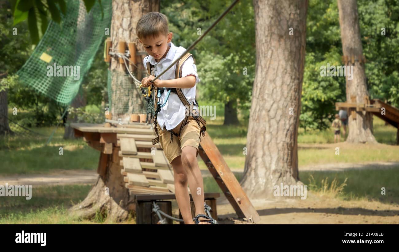 Little boy struggling walking on strung rope between trees in park ...