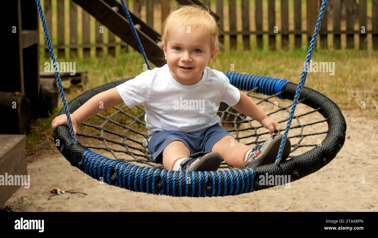 Cute smiling baby boy swinging in rope swing. Kids playing outdoors ...