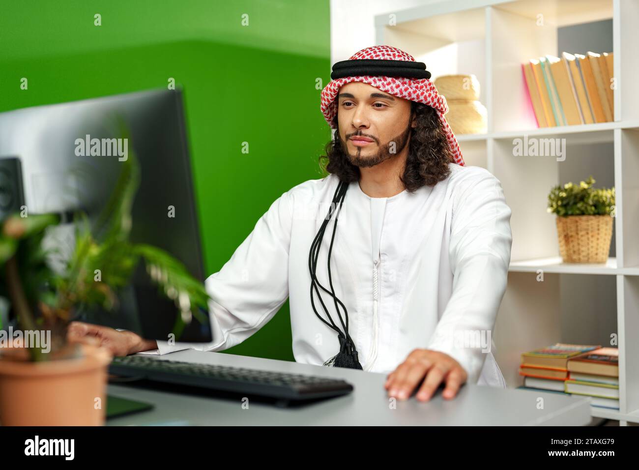 Young Arab man in traditional dress thobe working on computer in office ...