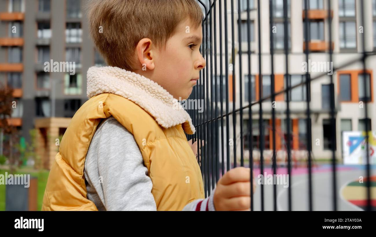 Sad little boy holding metal fence on public playground and looking through. Child depression ...