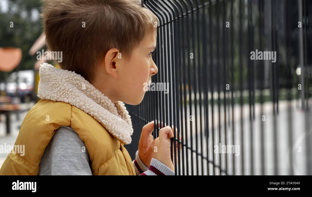 Little boy feeling sad and alone looking through metal fence on ...