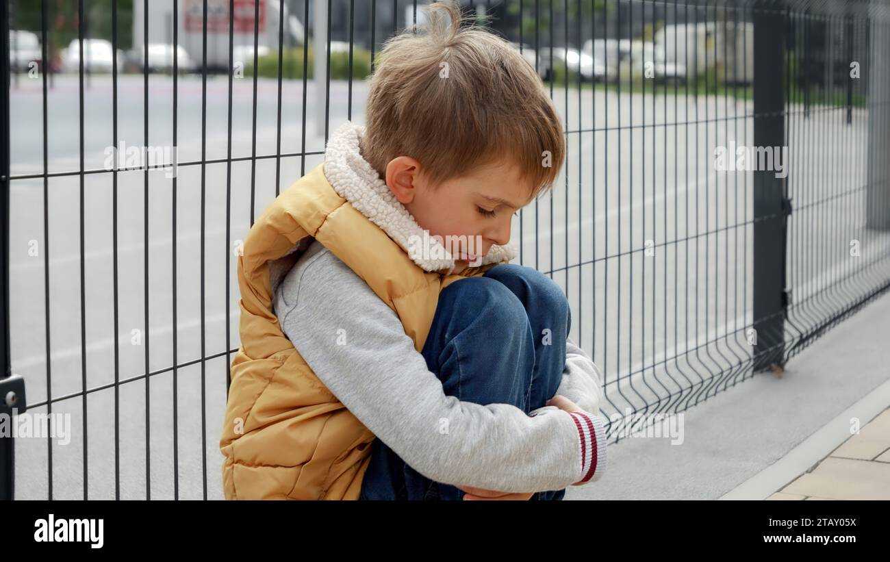 Portrait of little boy crying while sitting alone next to metal fence ...